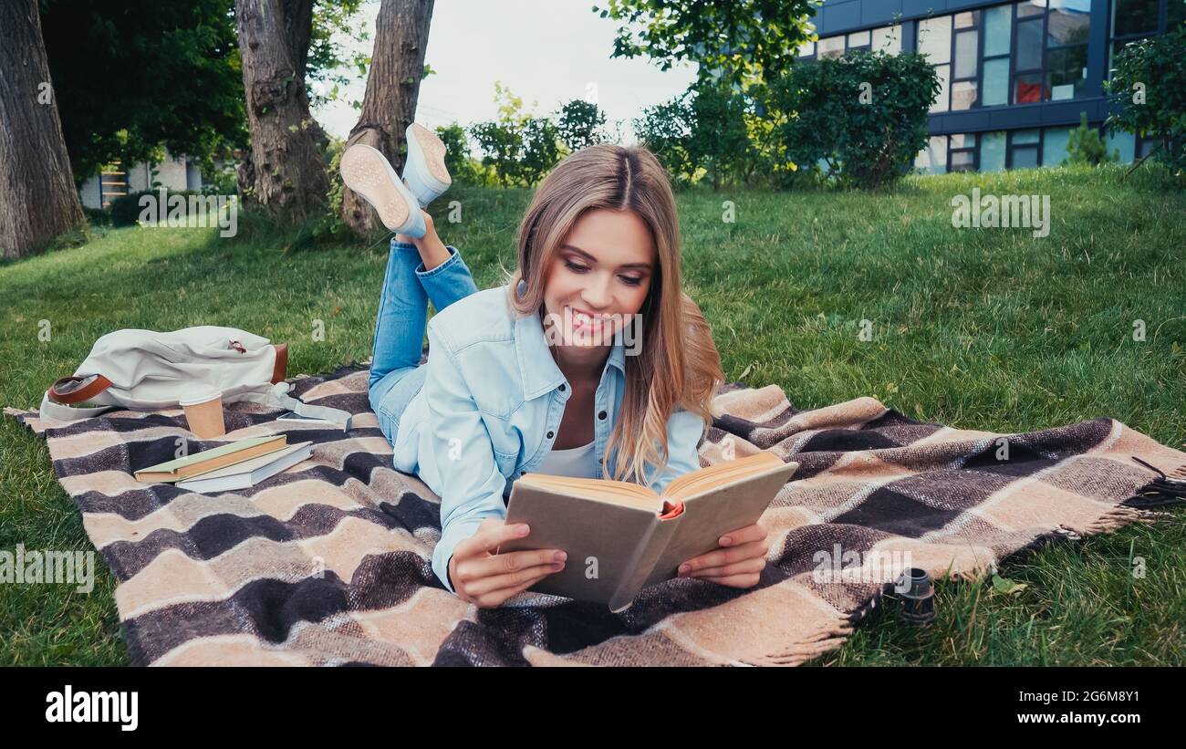 young cheerful student reading book while lying on checkered blanket in ...