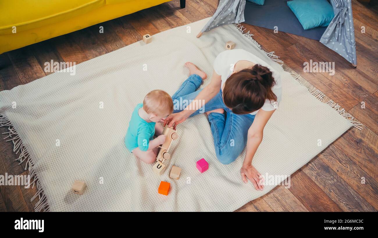 top view of barefoot mother sitting near infant son playing with toys ...
