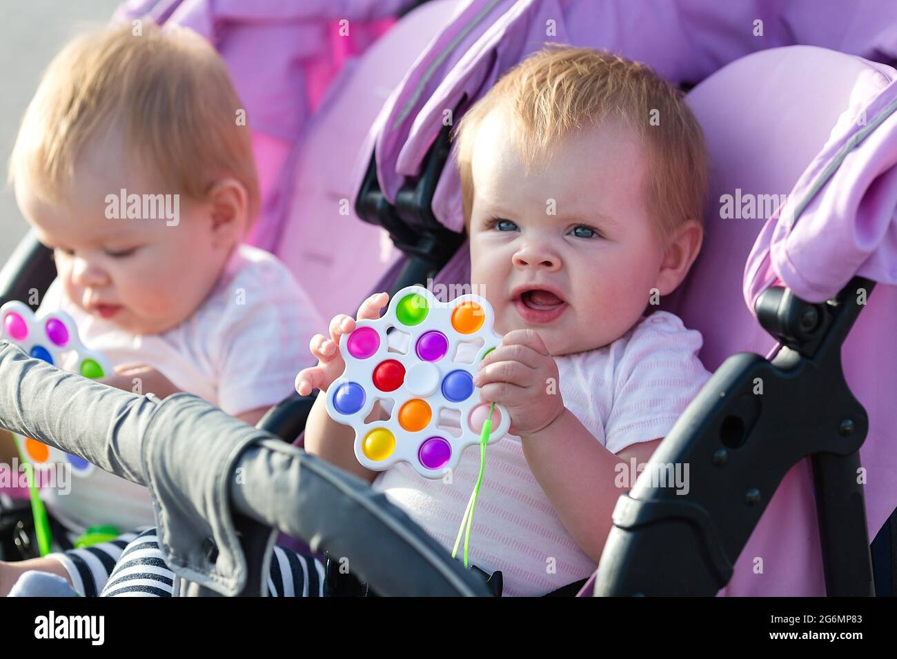 Smiling twin girls in a baby carriage with bright toys in their hand ...