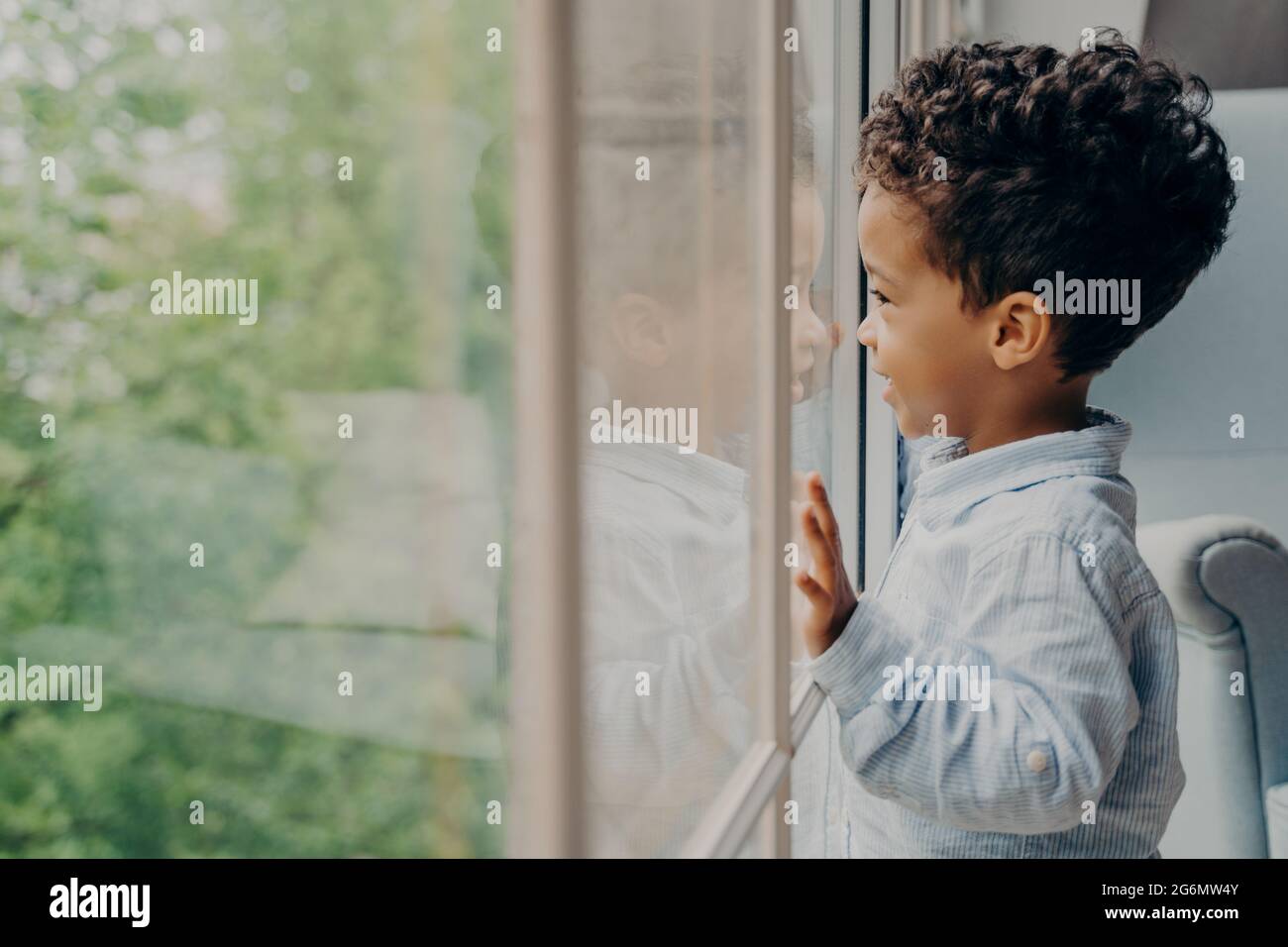 Lovely afro american kid in light blue shirt looking out of window ...