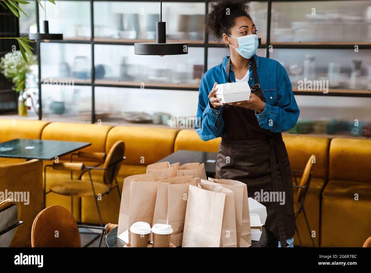 Black waitress in face mask holding food box while working at cafe ...