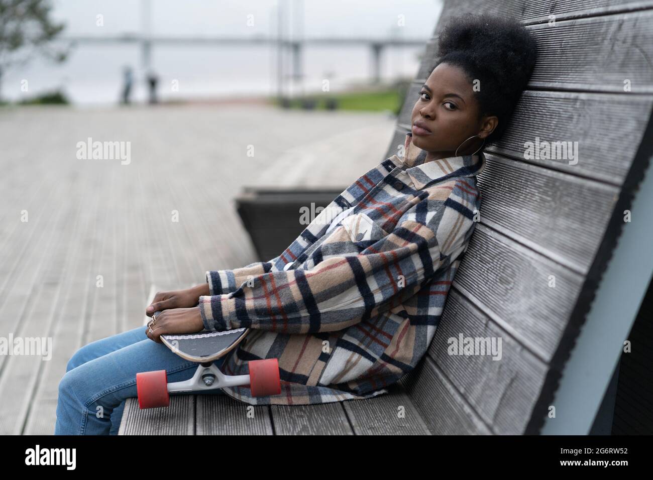 Grumpy african woman tired sit in urban space park with longboard look ...