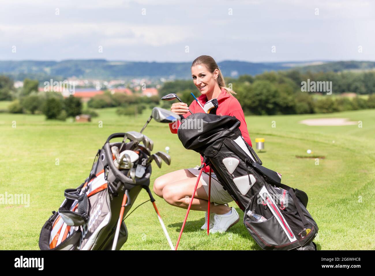 Portrait of smiling female player crouching choosing golf club from bag ...