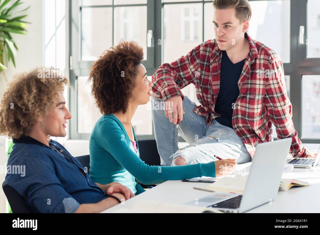 Relaxed young student sitting on desk while talking with his classmates ...