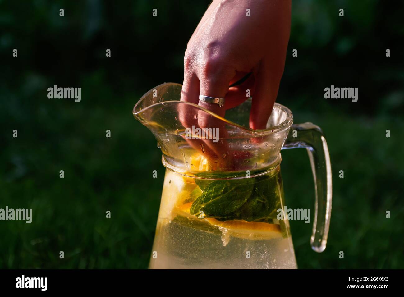 Defocus hand put strawberry in glass jug of lemonade with slice lemon and mint on natural deep ...