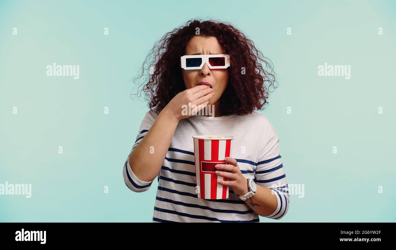 curly young woman in 3d glasses eating popcorn isolated on blue Stock ...