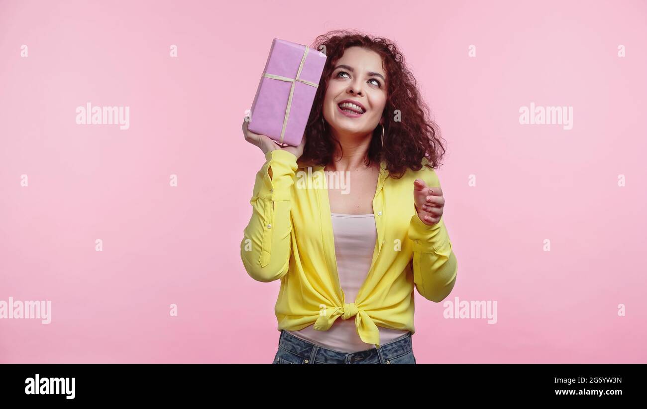 curious and smiling woman holding wrapped gift box isolated on pink ...