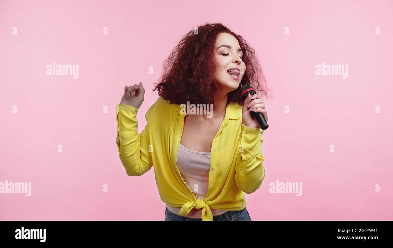 young curly woman sticking out tongue while singing in microphone ...