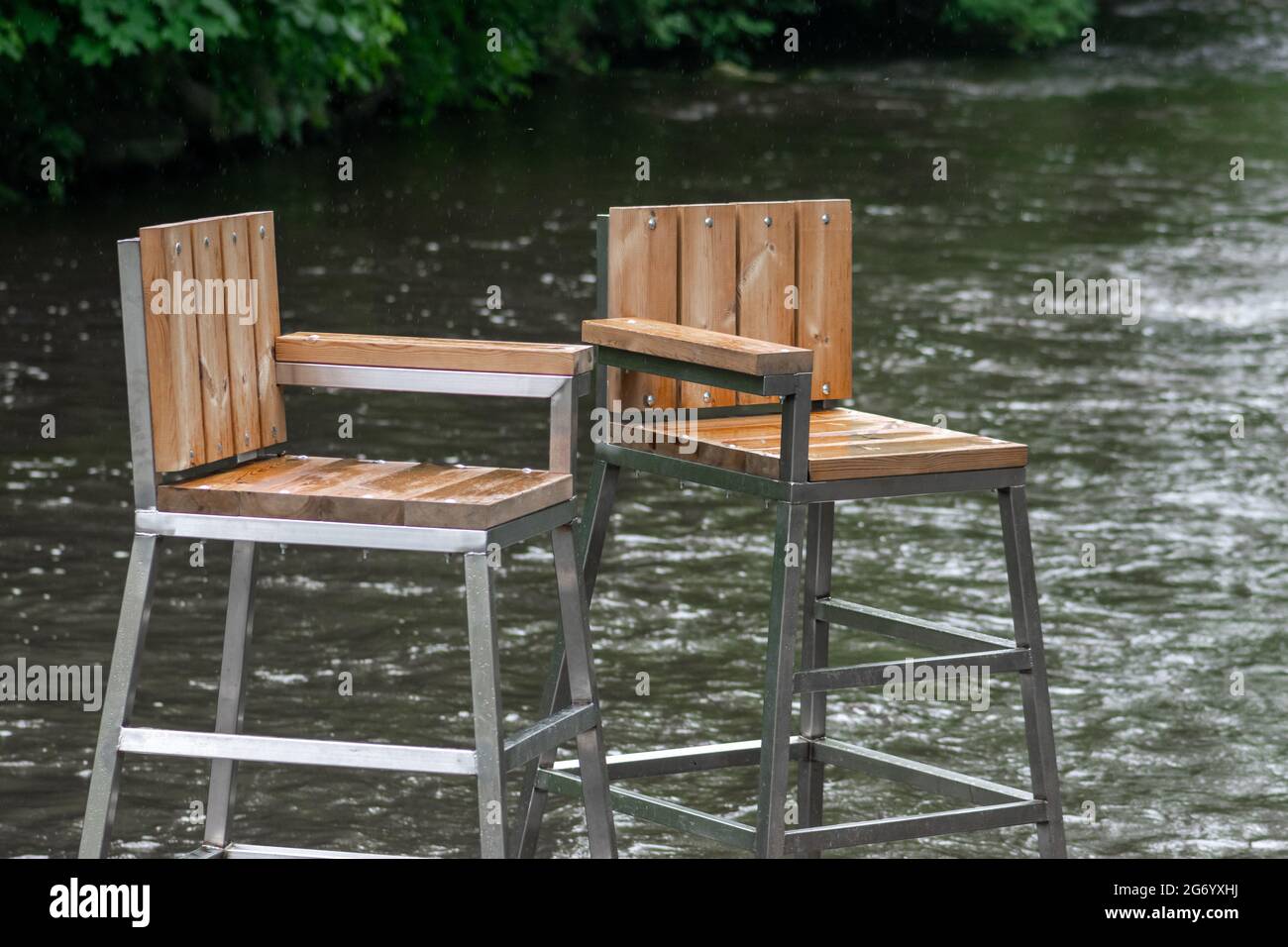 Two wooden chairs with metallic legs standing in river and green trees ...