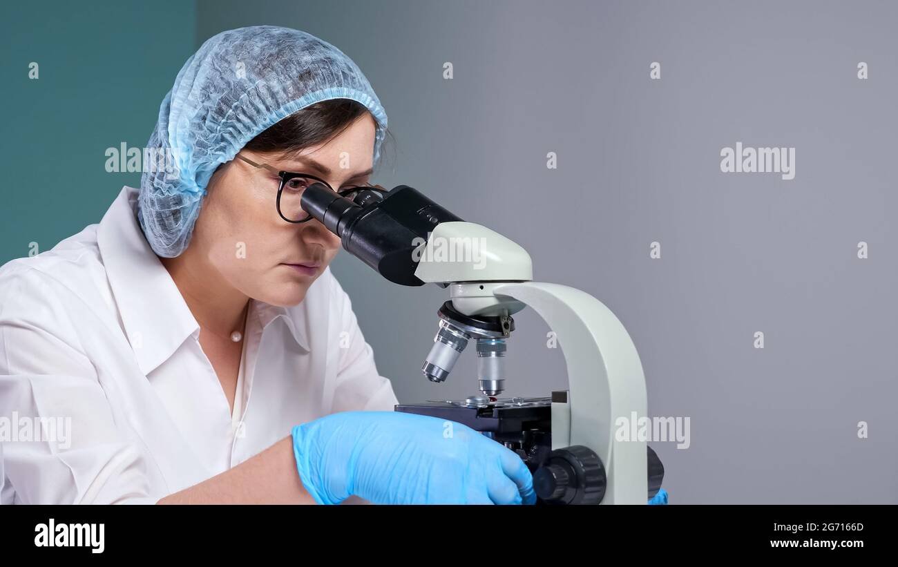 Female researcher in cap and white robe looks at blood sample through ...