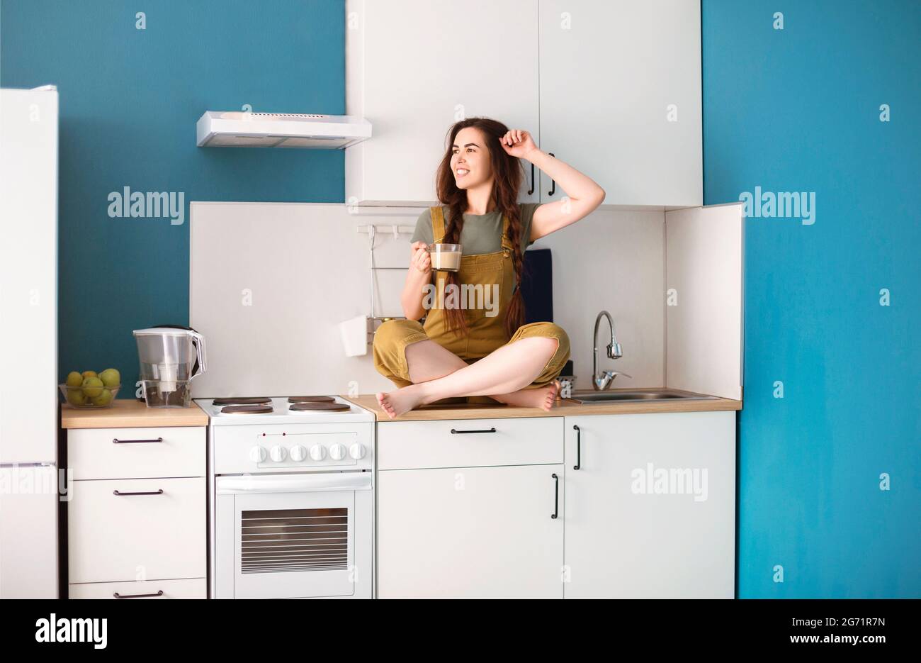Young barefoot female sitting on counter and enjoying coffee in modern ...