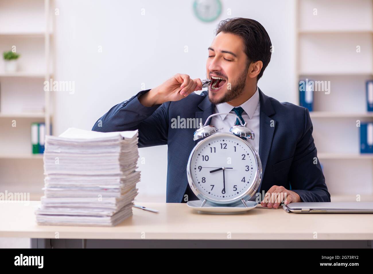 Young male employee eating alarm-clock Stock Photo - Alamy