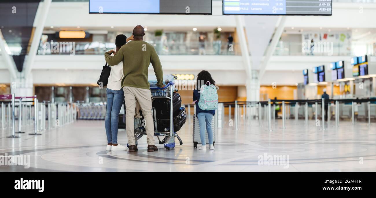 Rear view of family with luggage trolley waiting for flight at airport ...