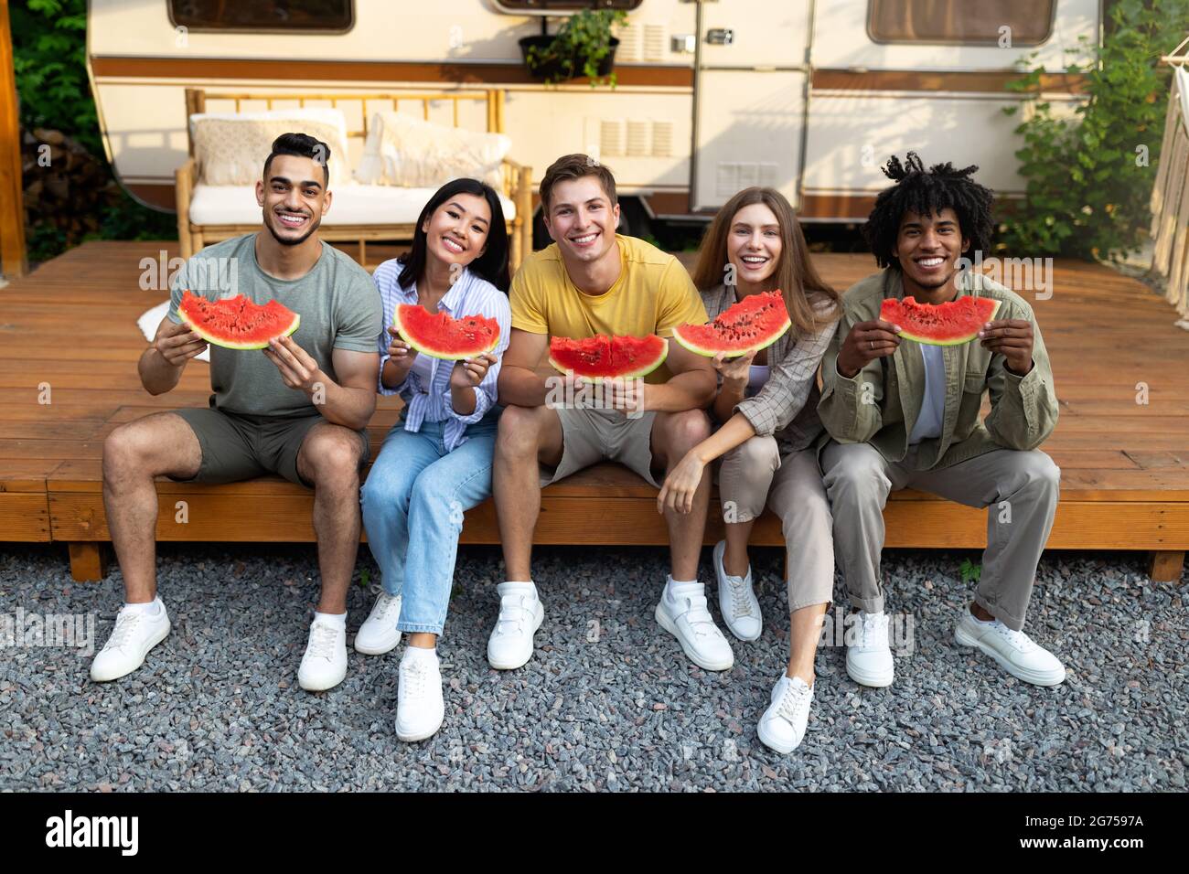 Happy diverse friends sitting near camper van with pieces of watermelon ...