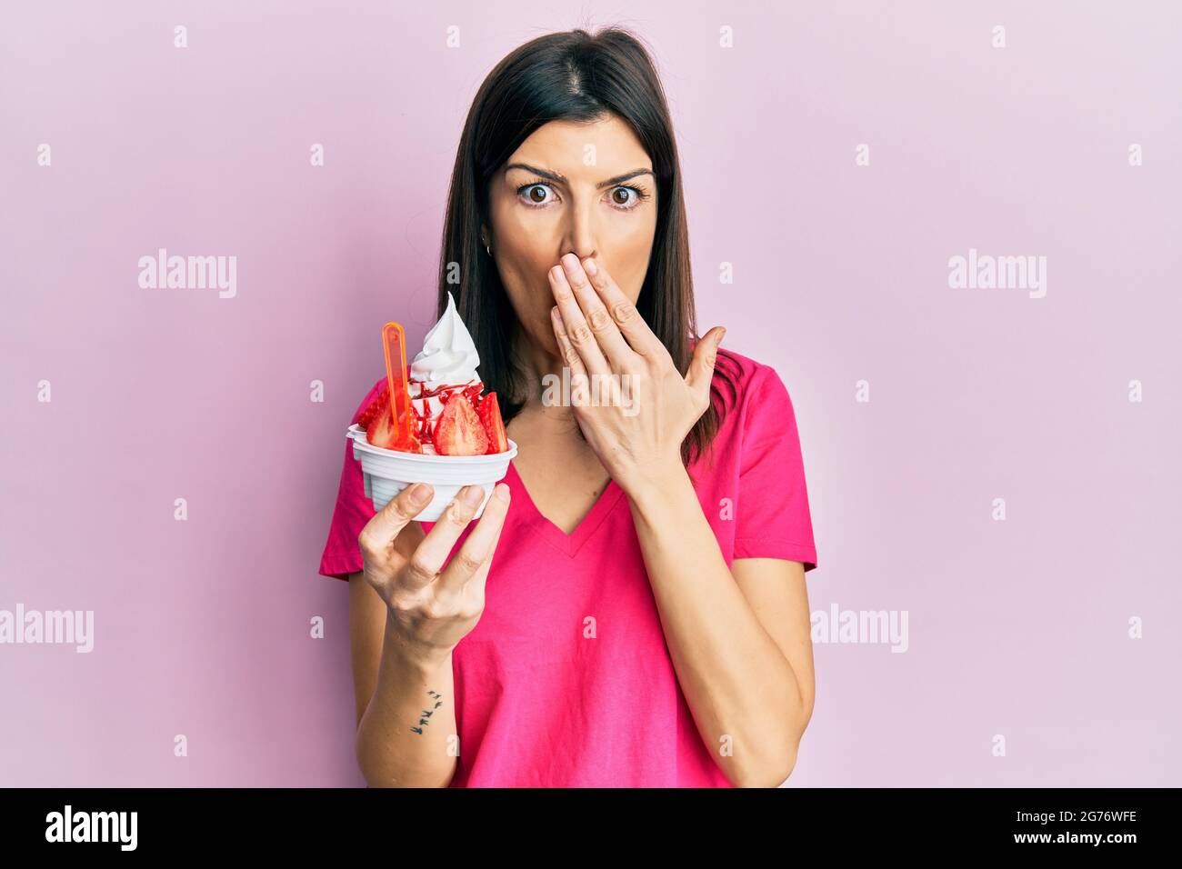 Young hispanic woman eating strawberry ice cream covering mouth with ...