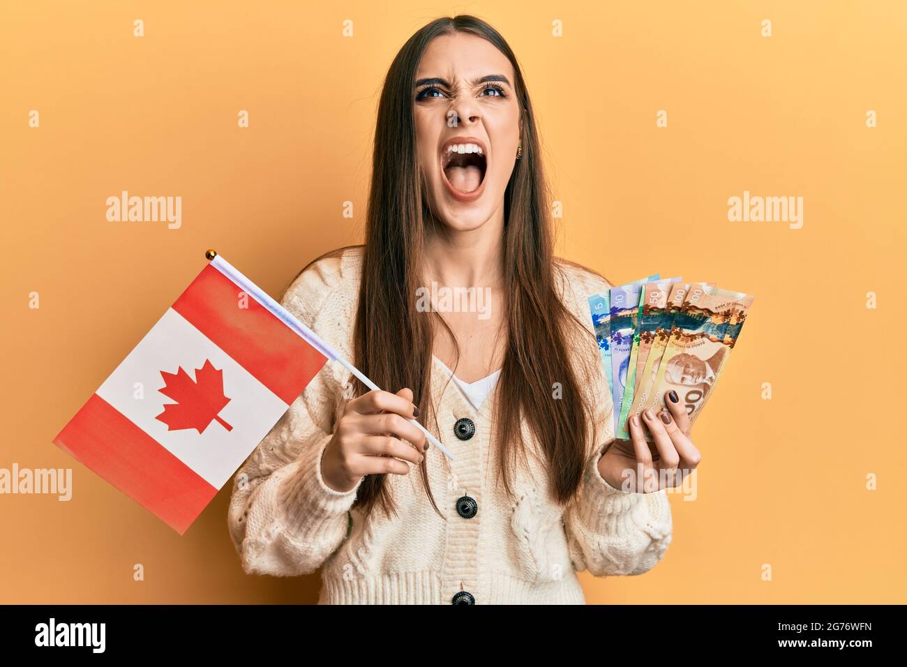 Beautiful brunette young woman holding canada flag and dollars angry ...