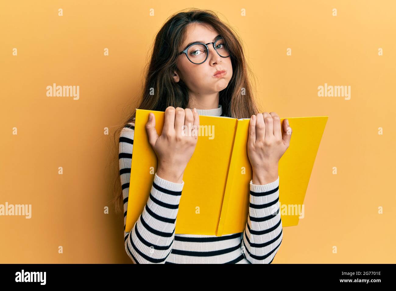 Young caucasian woman reading a book wearing glasses puffing cheeks with funny face. mouth ...