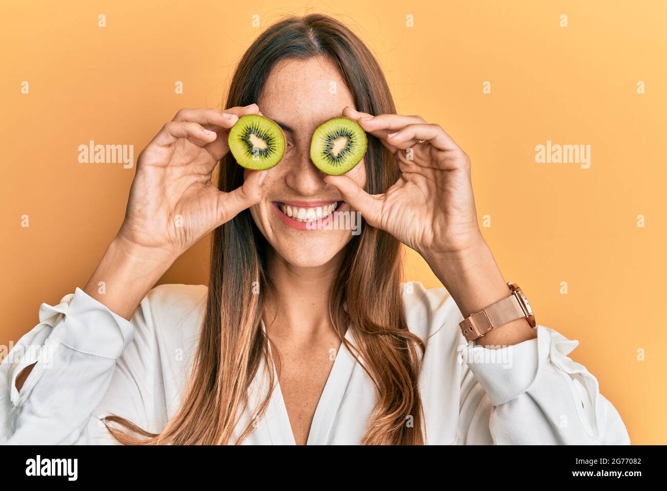 Young beautiful woman holding slice of kiwi over eyes smiling with a ...