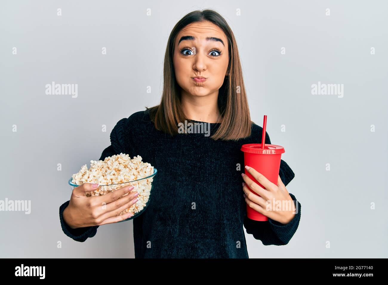 Young brunette girl eating popcorn and drinking soda puffing cheeks ...