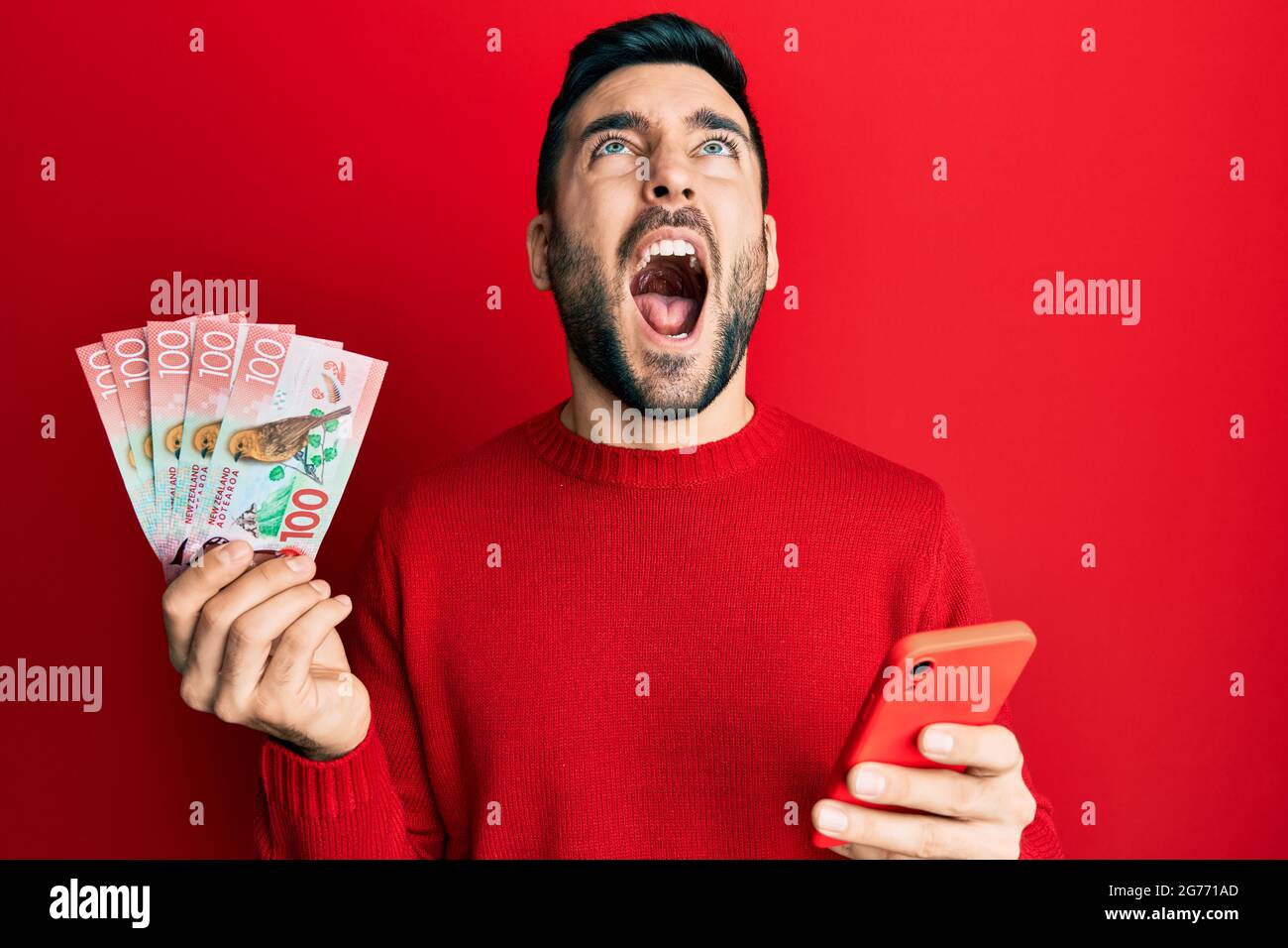 Young hispanic man using smartphone holding new zealand dollars ...