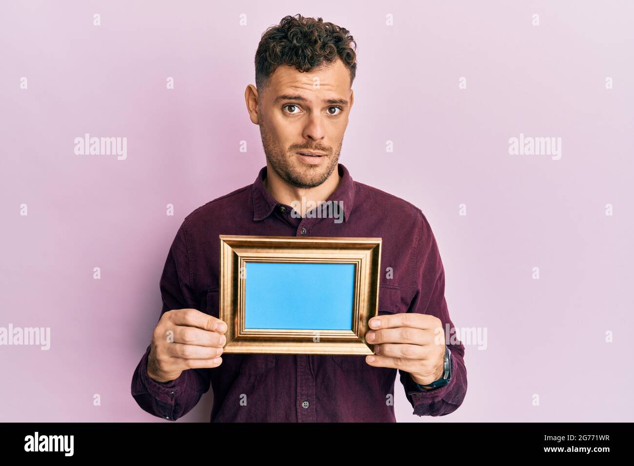 Young hispanic man holding empty frame in shock face, looking skeptical ...