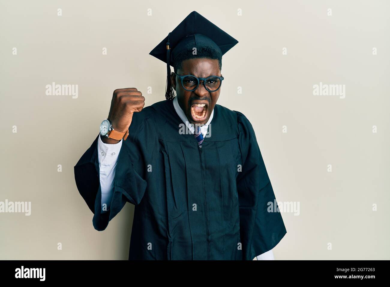 Handsome black man wearing graduation cap and ceremony robe angry and ...