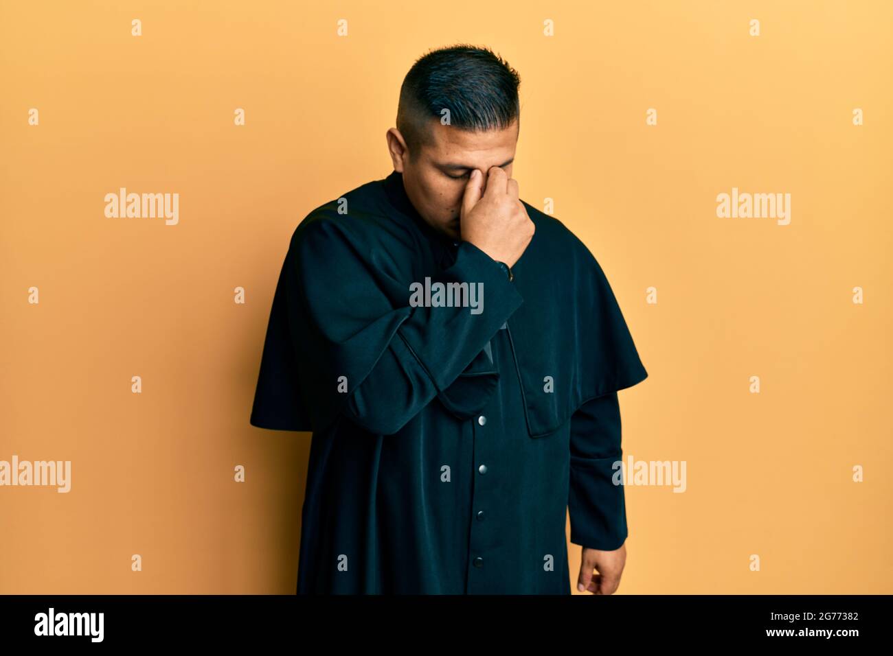 Young latin priest man standing over yellow background tired rubbing ...