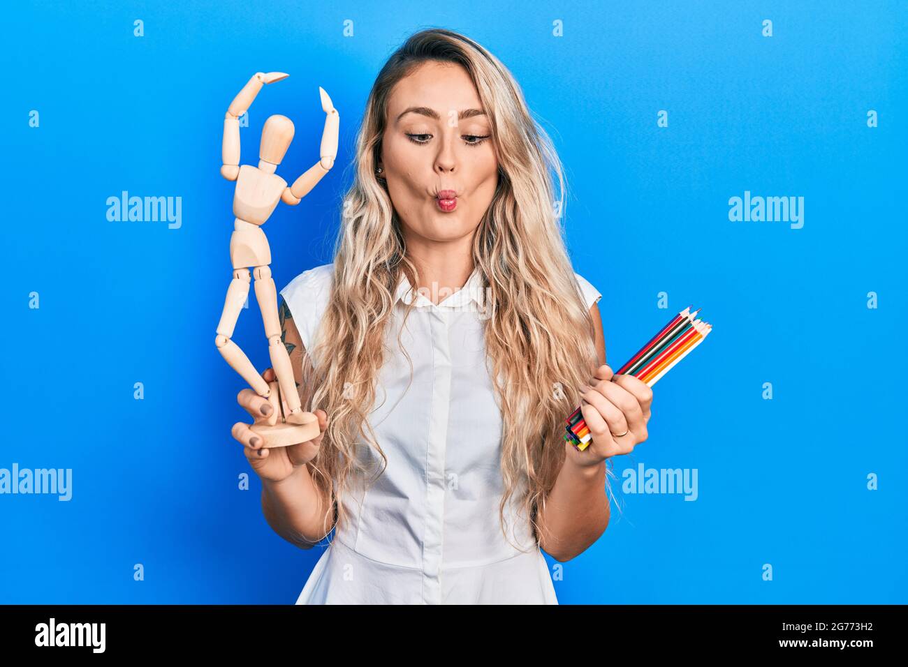 Beautiful young blonde woman holding wooden manikin and colored pencils ...