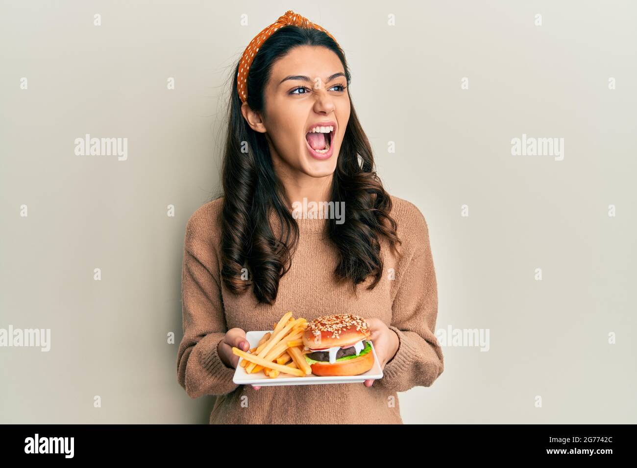 Young hispanic woman eating a tasty classic burger angry and mad ...