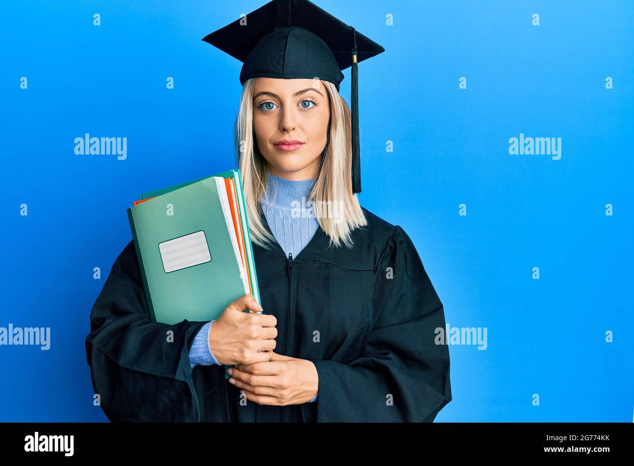 Beautiful blonde woman wearing graduation cap and ceremony robe holding ...