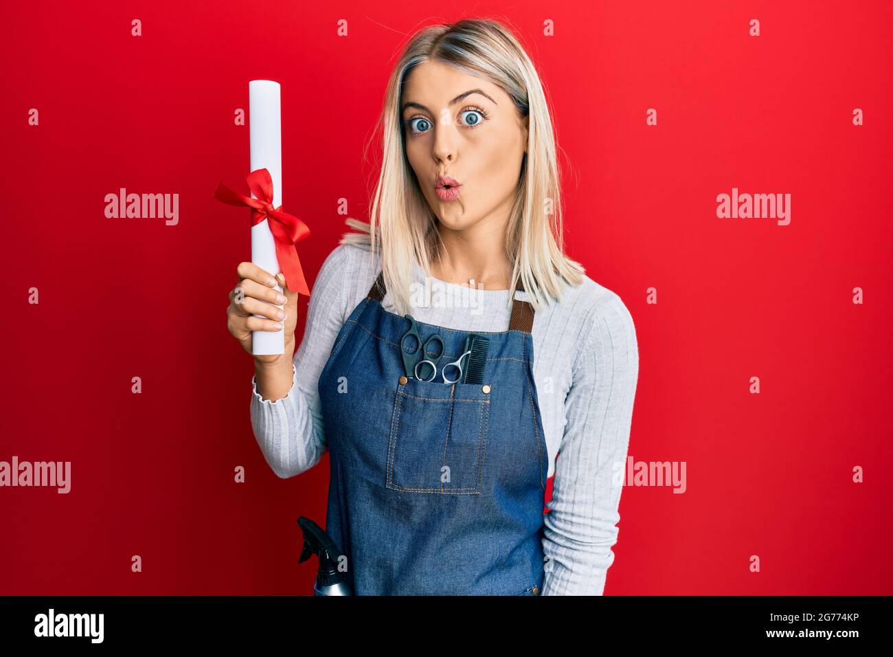 Beautiful blonde woman wearing hairdresser apron holding degree scared and amazed with open ...