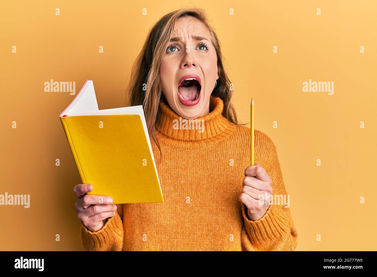 Young blonde woman holding book and pencil angry and mad screaming ...