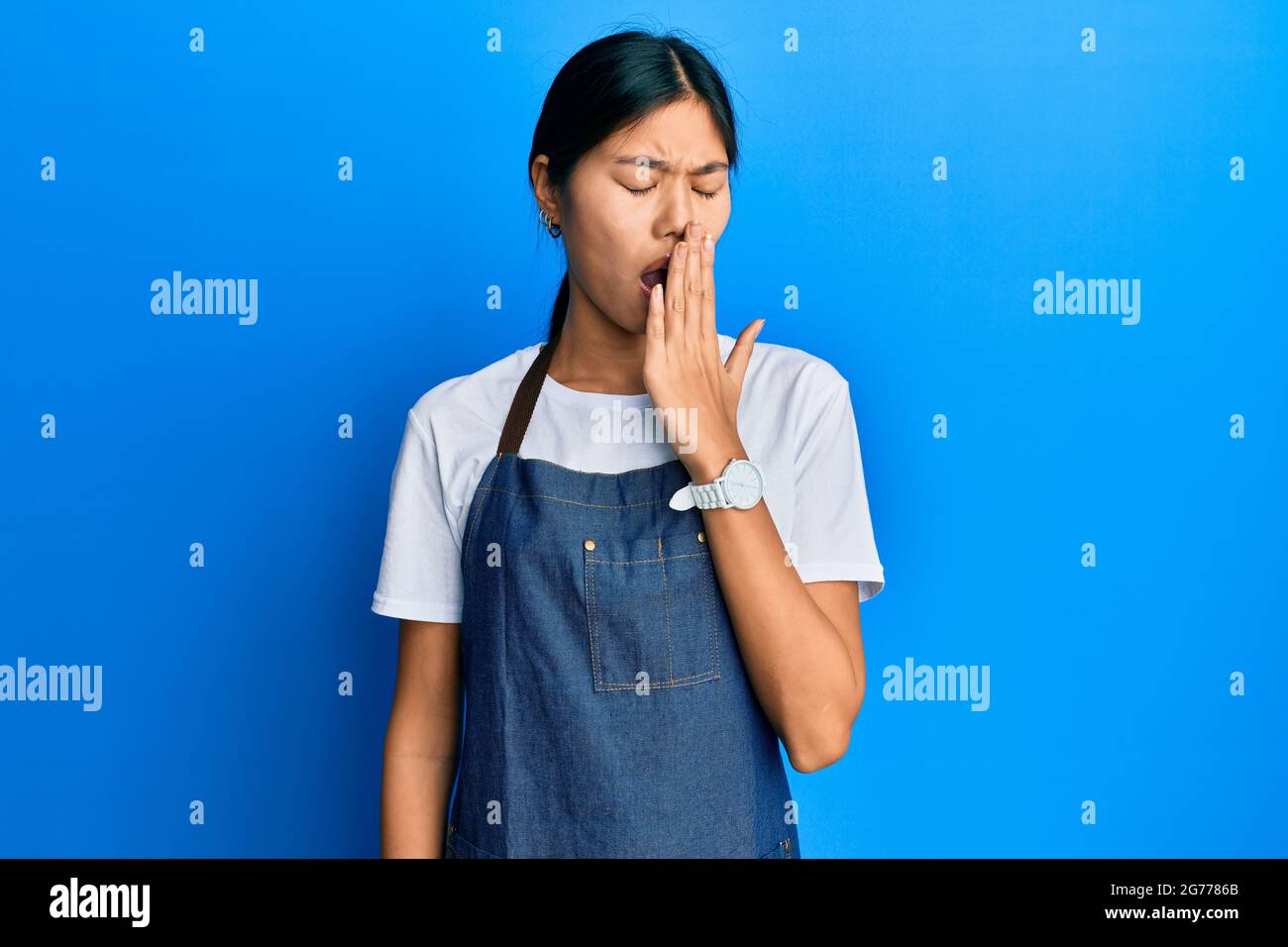 Young chinese woman wearing waiter apron bored yawning tired covering ...