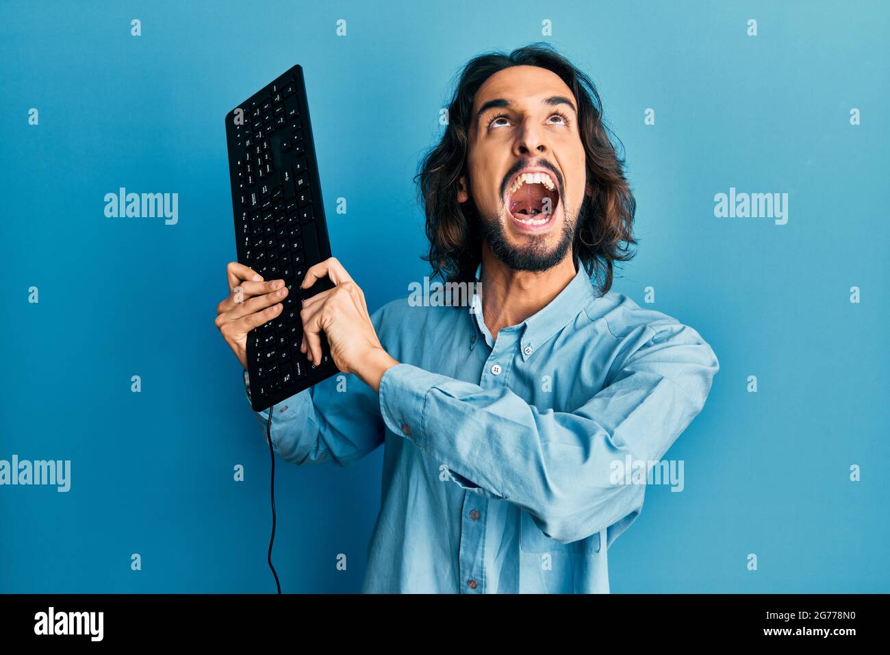 Young hispanic man holding keyboard angry and mad screaming frustrated ...