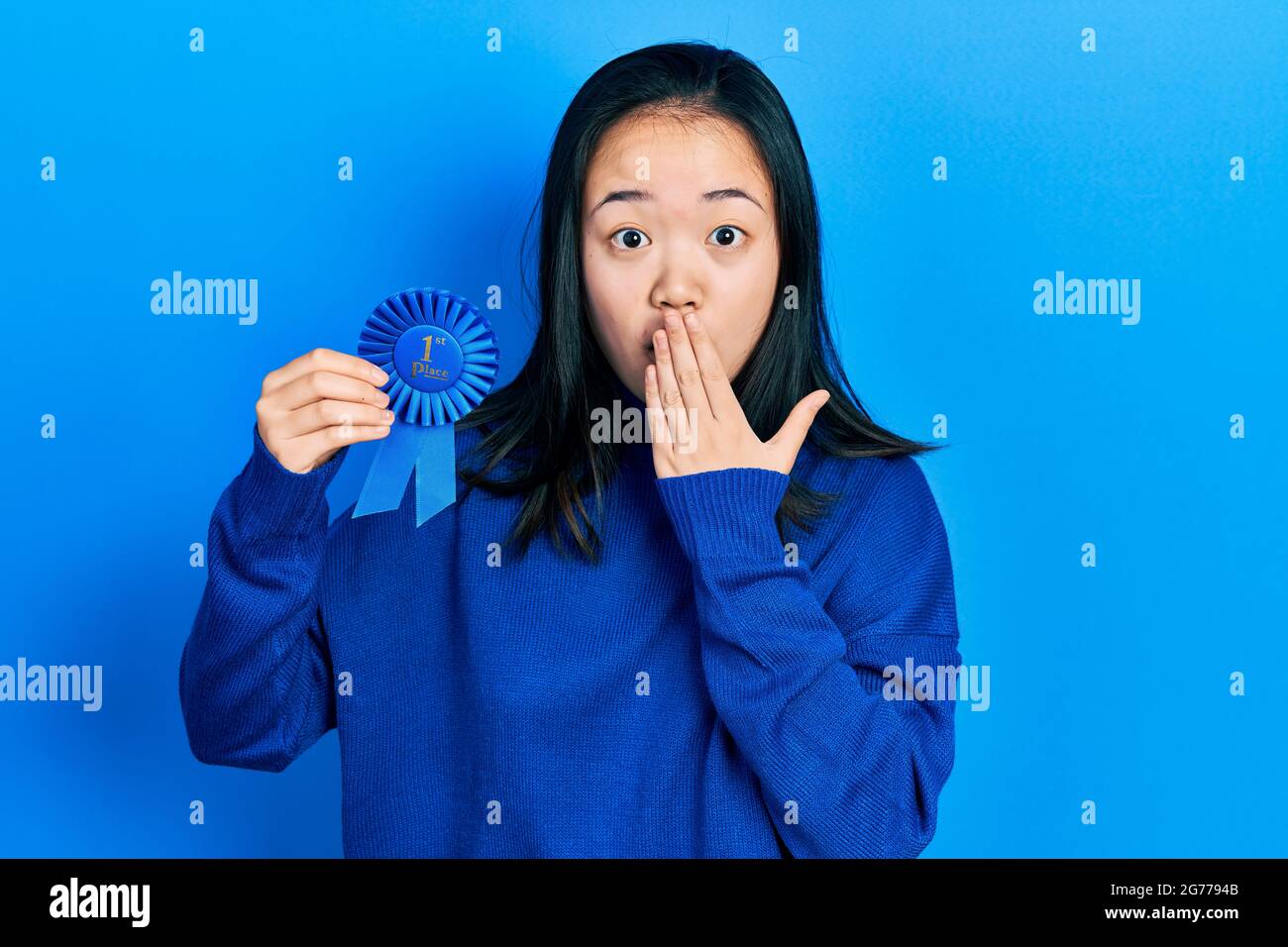 Young chinese girl holding first place badge covering mouth with hand ...