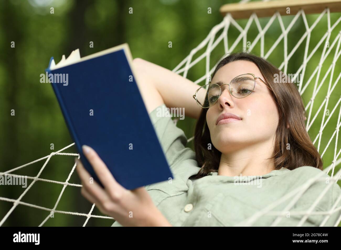 Relaxed woman reading a paper book wearing eyeglasses on hammock in a ...