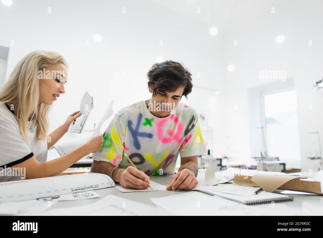blonde designer holding sewing patterns near colleague drawing with ...