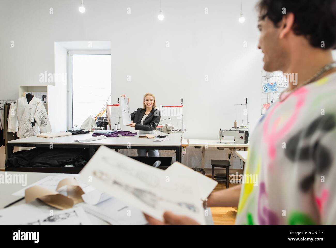 smiling fashion designer showing sewing pattern to colleague on blurred ...