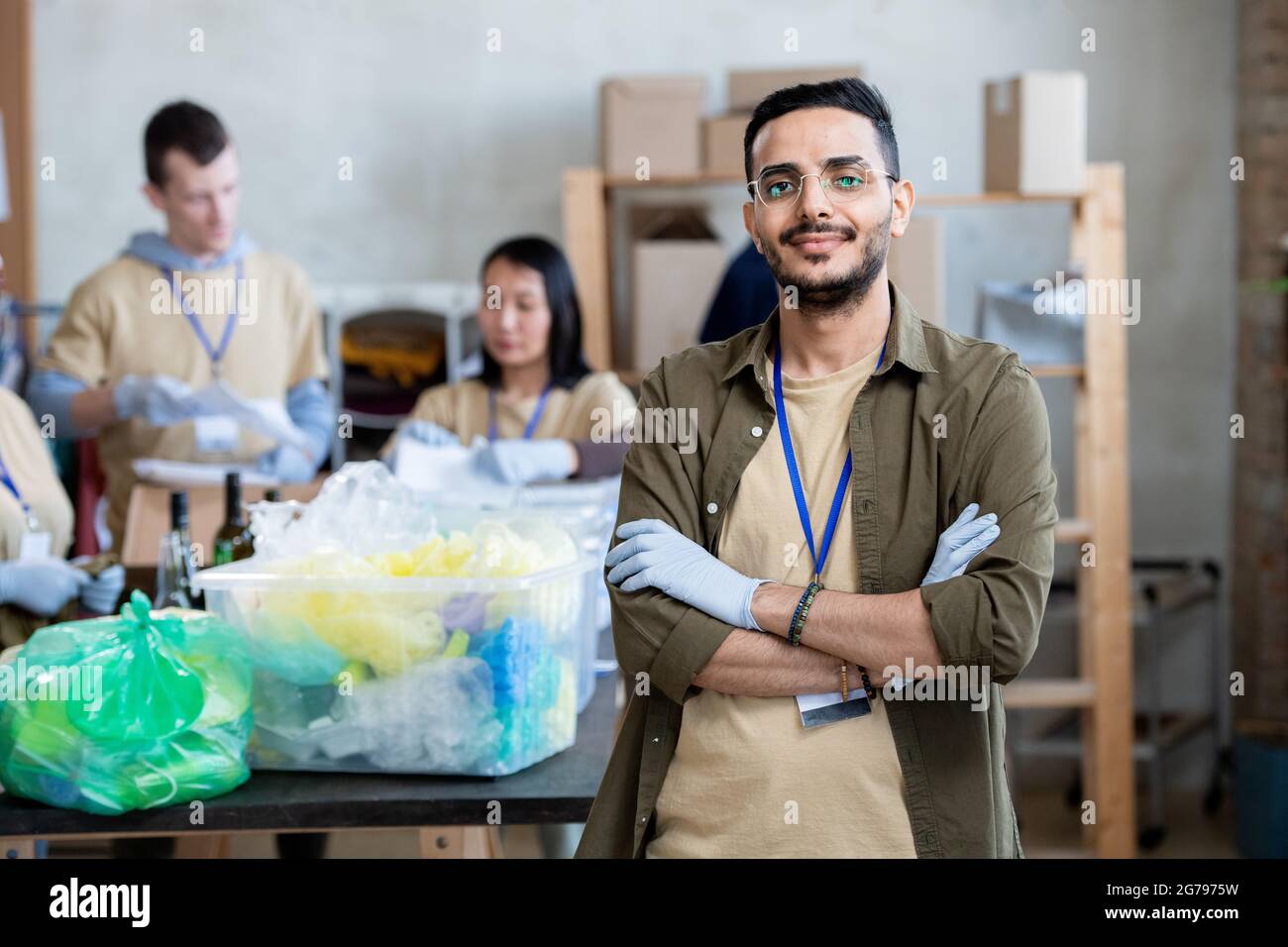 Gloved young man crossing arms by chest against volunteers sorting ...