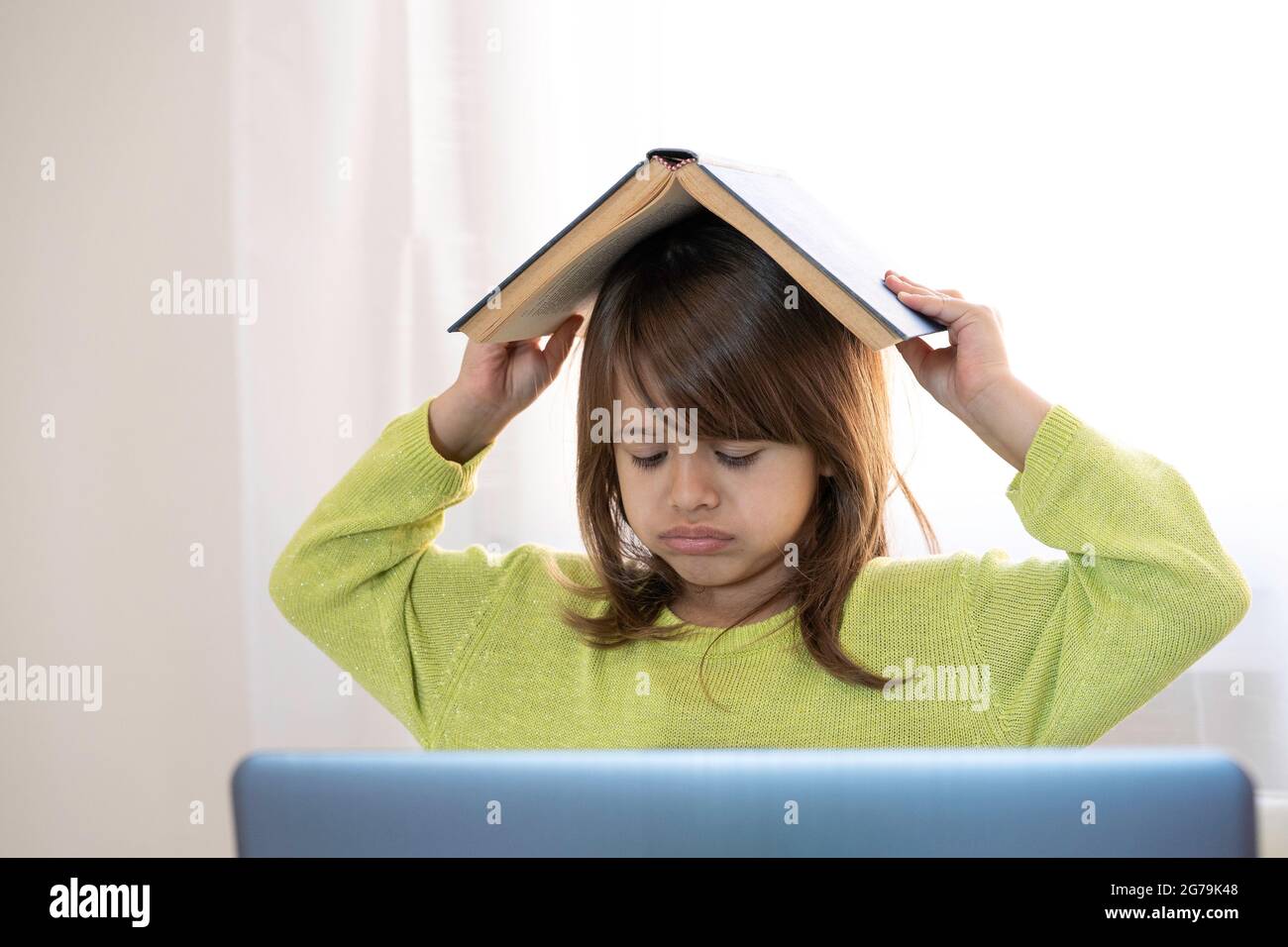 Sad elementary schooler girl studying at home with a book on her head ...