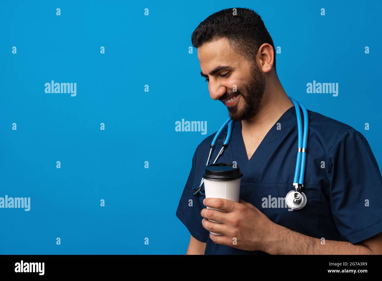 Young arab doctor drinking cup of coffee standing over blue background ...