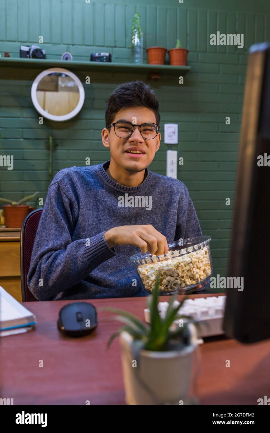 Young smiling Hispanic male sitting in front of the computer and eating ...