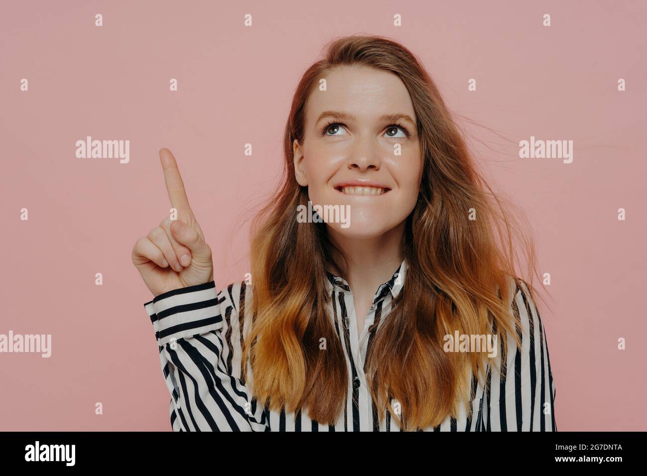 Young woman thinking while biting lips and having pointer finger up ...