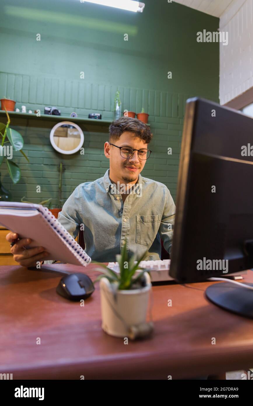 Young smiling Hispanic male working on a computer holding a notebook ...