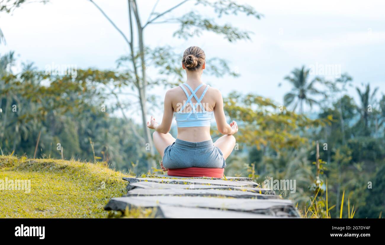 Young woman on path into the tropical jungle in yoga lotus pose doing ...