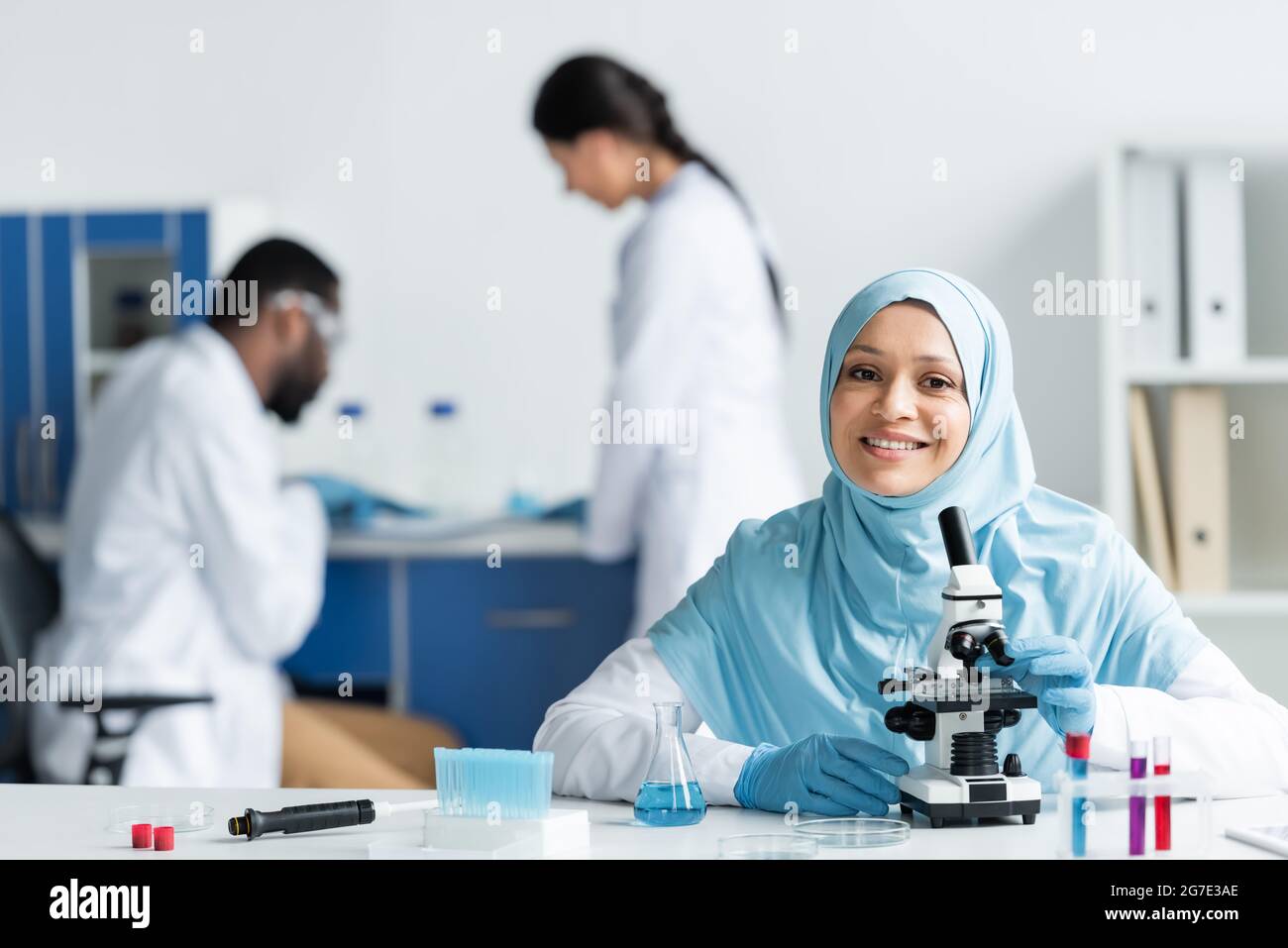 Positive muslim scientist looking at camera near microscope, flask and ...