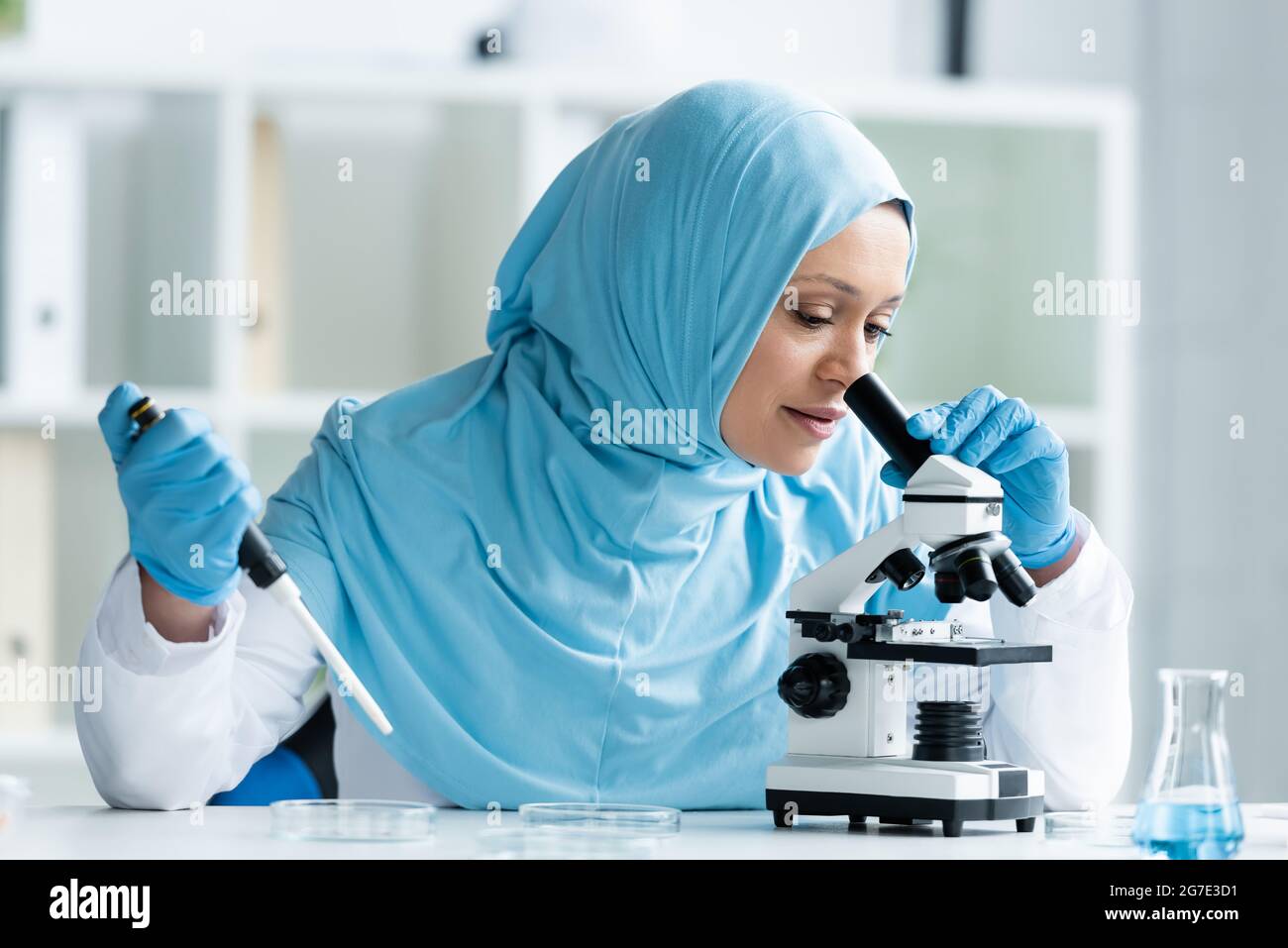 Arabian scientist in hijab looking through microscope and holding ...