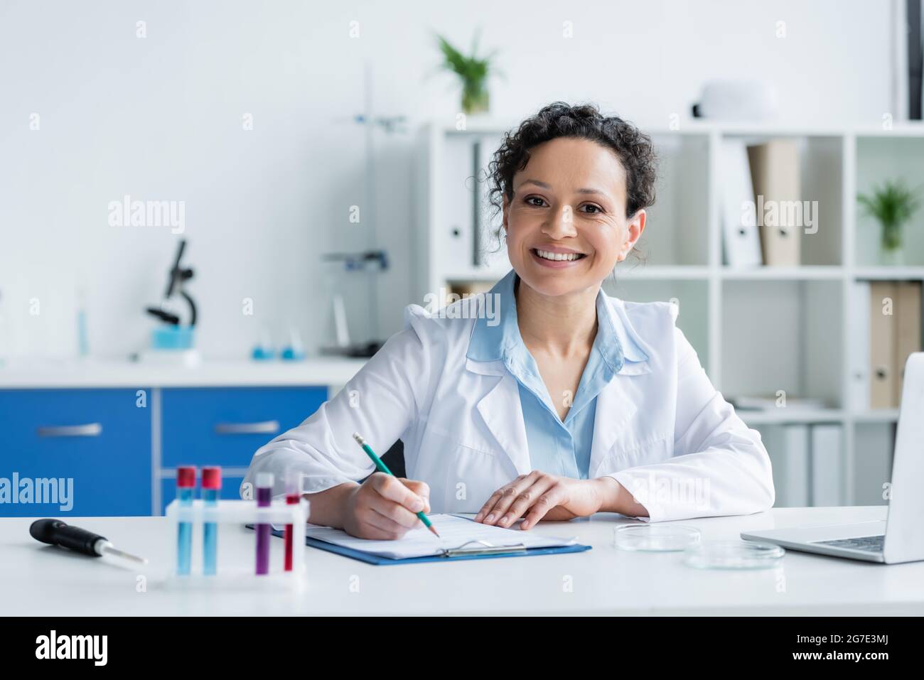 Smiling african american scientist writing on clipboard near blurred ...