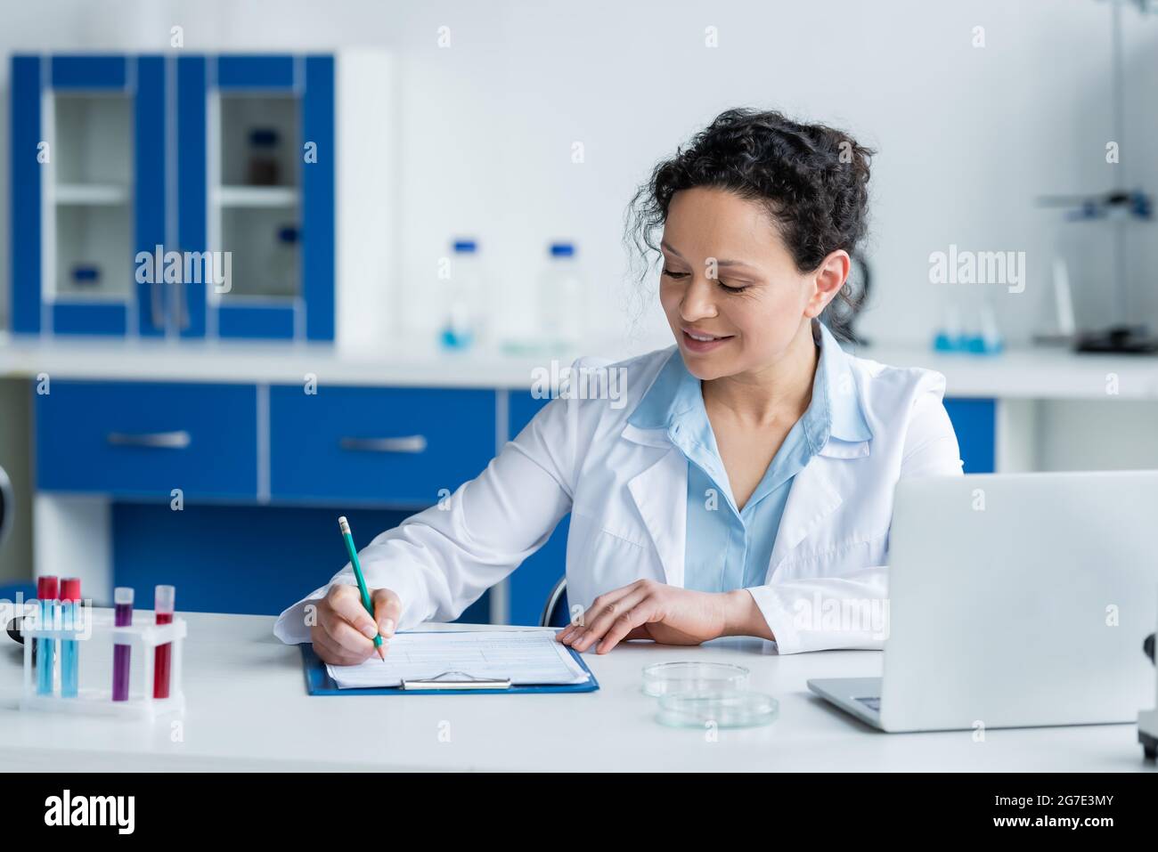 Smiling african american scientist writing on clipboard near laptop and ...