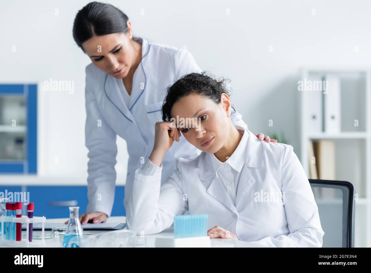 Sad african american scientist sitting near test tubes and colleague on ...